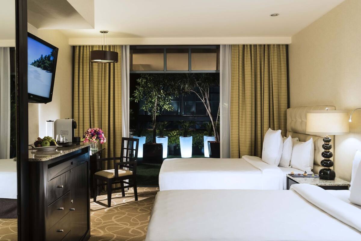 Quarto de hotel com duas camas, uma mesa de escritório com um computador, televisão, uma cadeira e uma janela com cortinas.