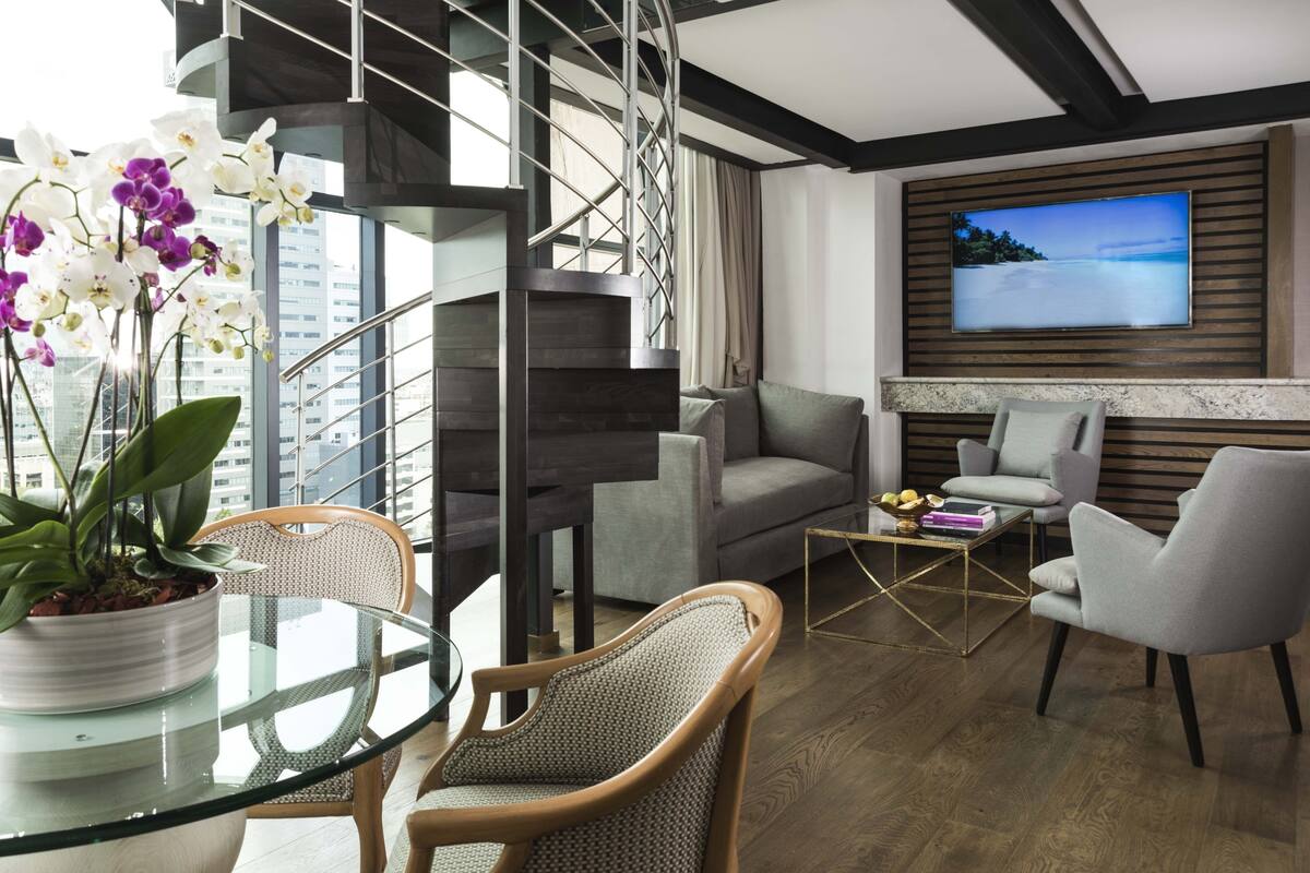 Uma sala de estar moderna com uma mesa de jantar de vidro, cadeiras de vime, um sofá cinza e uma televisão de tela plana fixada na parede.
