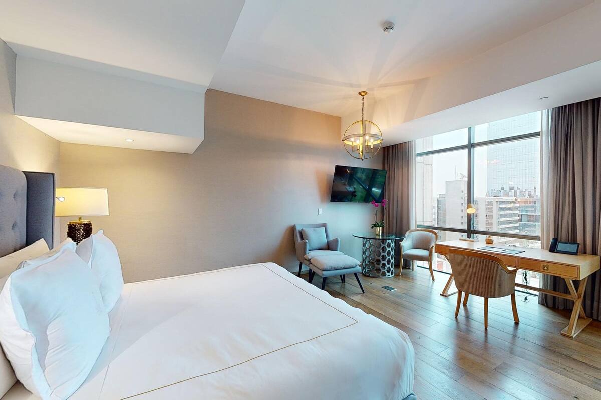 Quarto de hotel com uma cama grande, uma escrivaninha, uma cadeira, uma televisão e vista para a cidade através de grandes janelas.