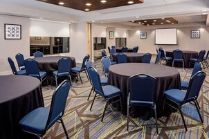 Sala de conferências com mesas redondas e cadeiras azuis, piso coberto por carpete e tela de projeção.