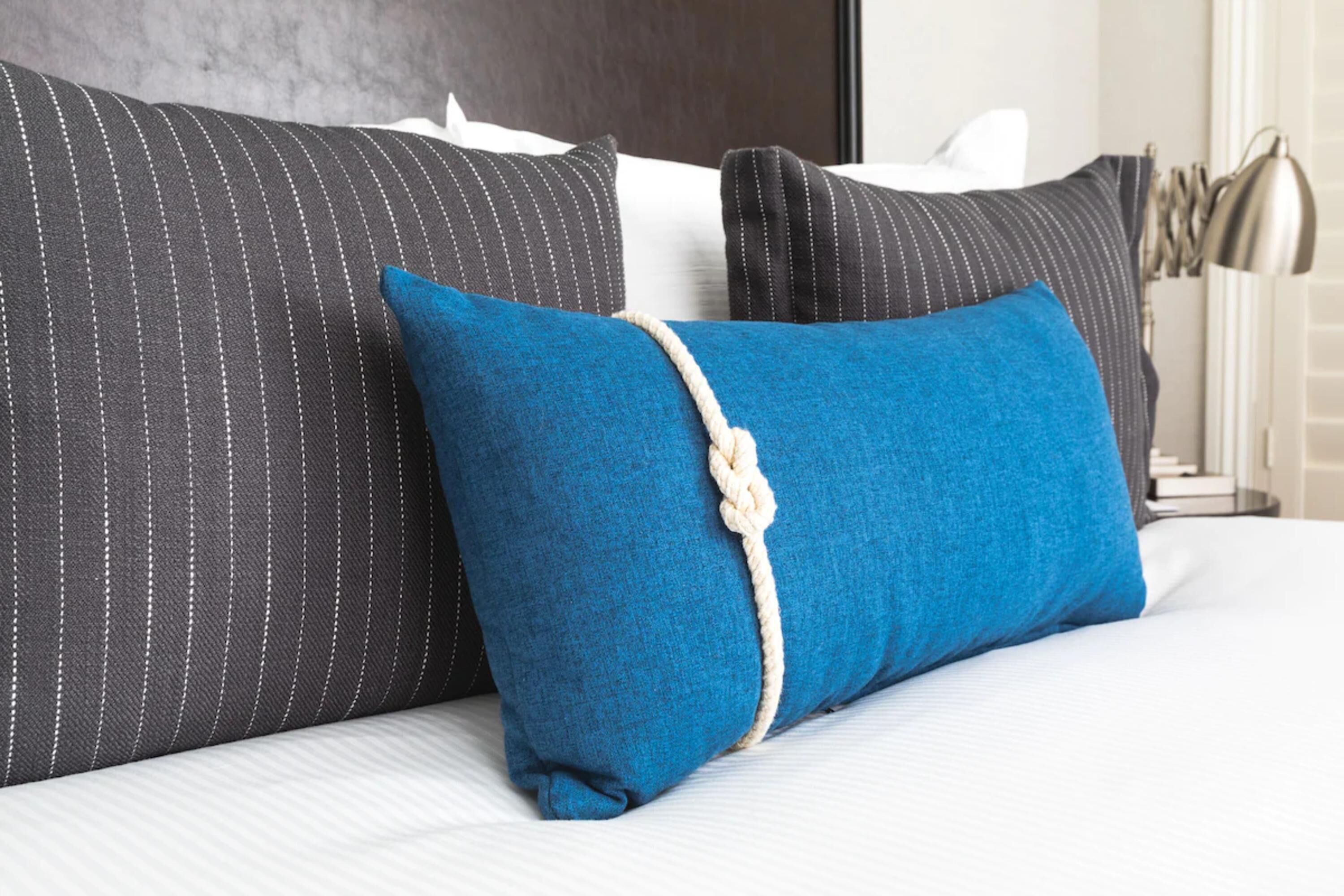 Cama com almofadas listradas e uma almofada azul com detalhe de corda.