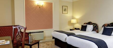 Quarto de hotel com duas camas, uma cadeira de madeira, uma mesa com abajur e um quadro na parede.