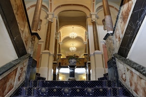 Uma escadaria imponente com colunas de mármore e um candelabro, que leva a um espaço amplo com pé-direito alto e um vaso grande sobre um pedestal.