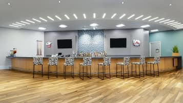 Uma área de bar no hall com balcão curvo, vários bancos altos, uma TV na parede e a placa 'Lobby Bar'.