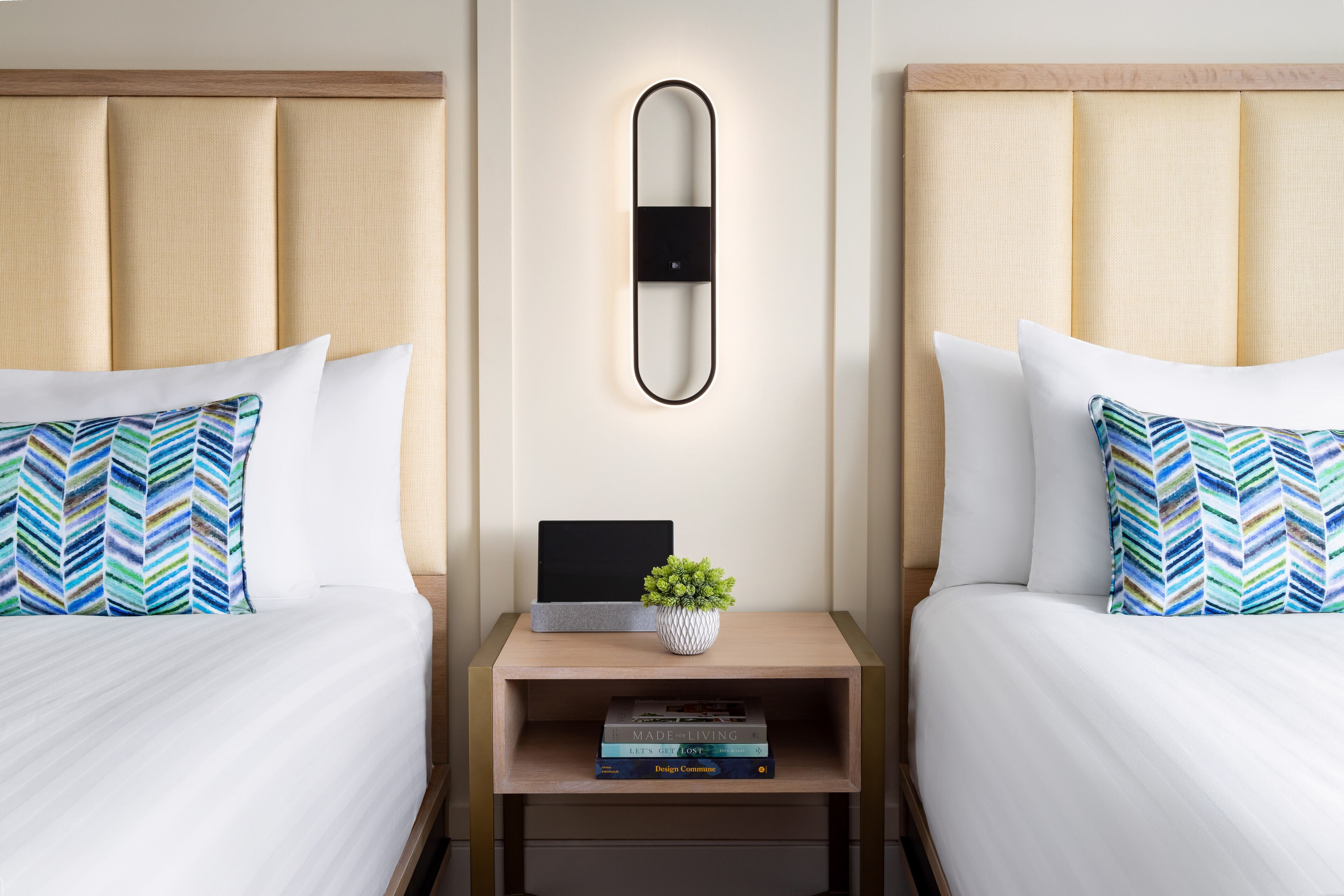 Quarto de hotel com duas camas, cada uma com edredom branco e almofadas estampadas. Há uma mesinha de cabeceira com uma pequena planta e alguns livros.