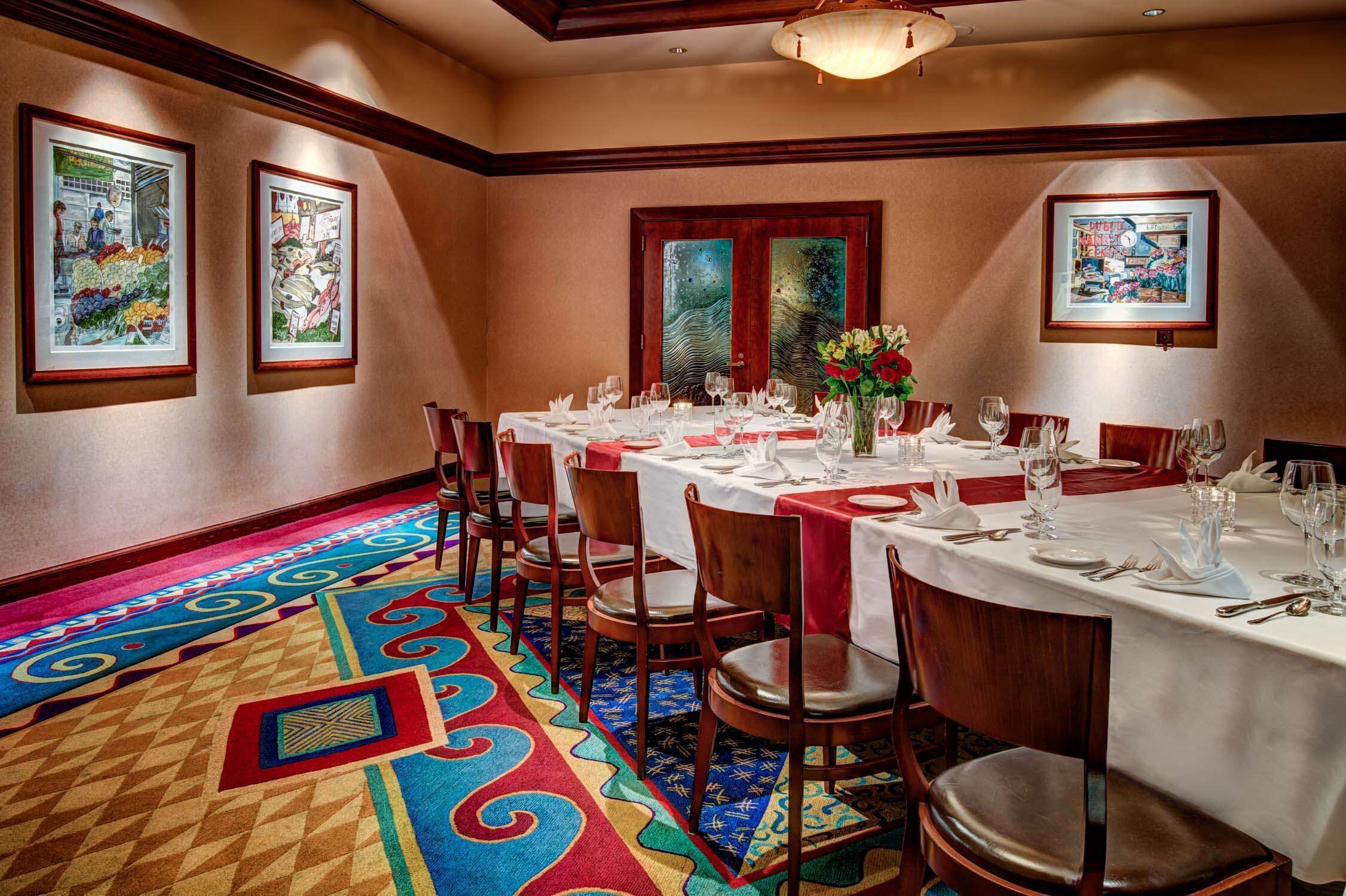 Uma sala de jantar com uma mesa comprida posta para refeição, obras de arte emolduradas nas paredes e um tapete colorido.