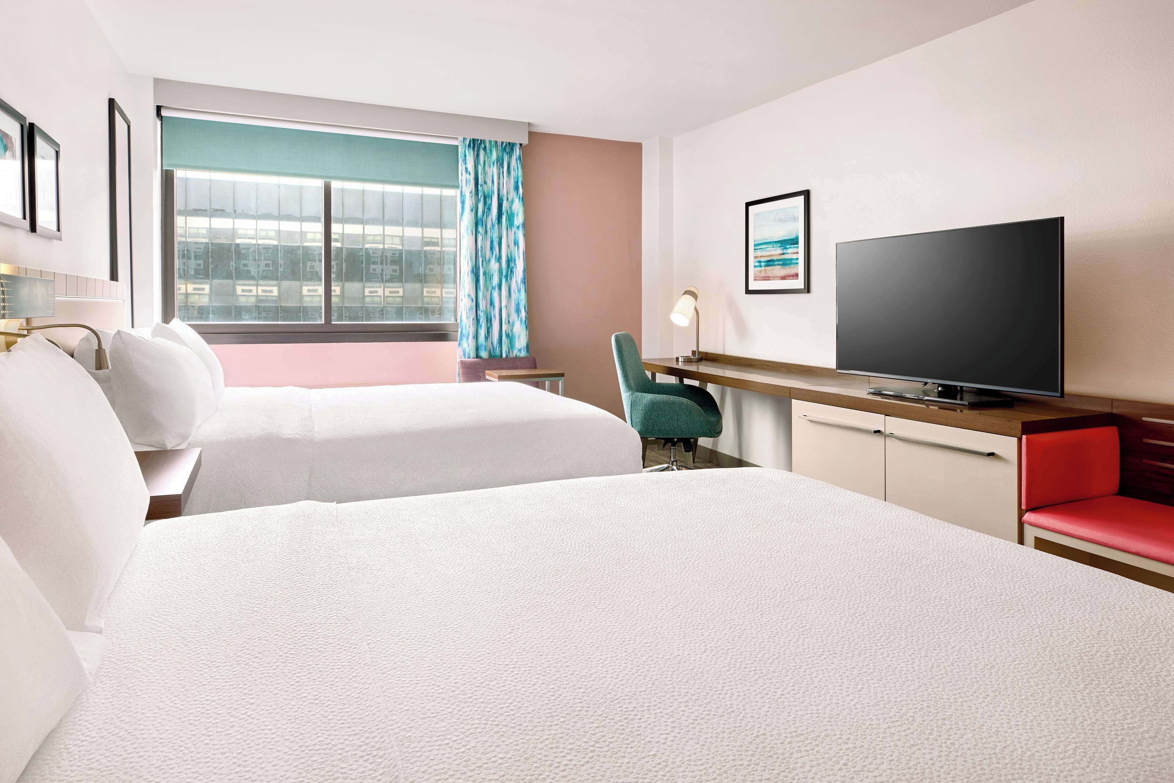 Quarto de hotel com duas camas, uma escrivaninha com televisão, uma cadeira e janela com vista para prédios.