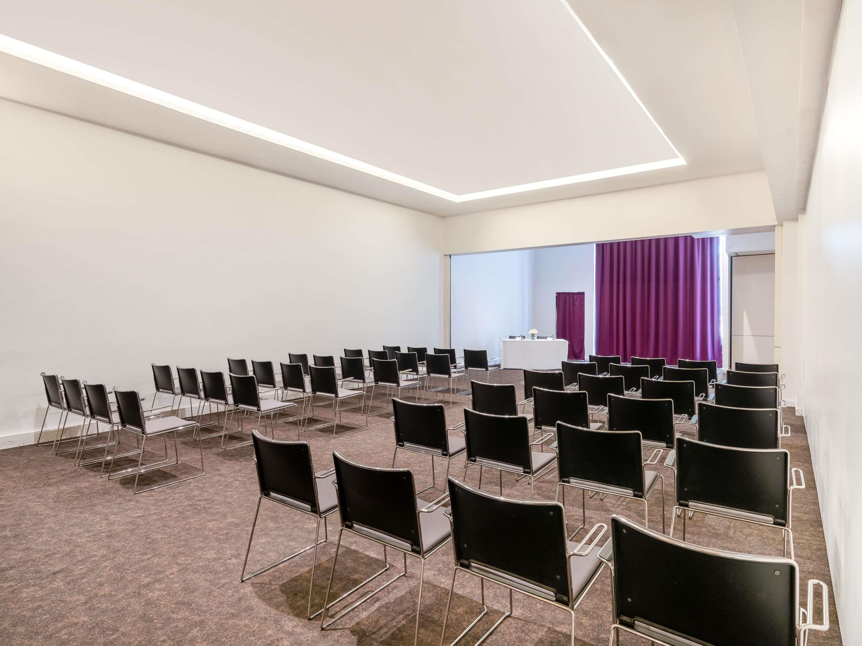 Uma sala de conferências com fileiras de cadeiras, uma mesa branca com um laptop e uma cortina roxa.