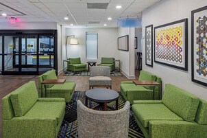 Um saguão de hotel com assentos estofados em tons de verde, uma mesa de centro e obras de arte nas paredes.