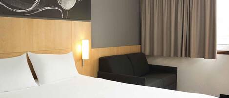 Quarto de hotel com uma cama grande, uma cadeira, uma luminária na parede e uma janela com cortinas.