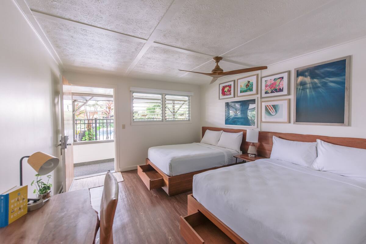 Quarto com duas camas, uma escrivaninha de madeira, um ventilador de teto e obras de arte nas paredes.