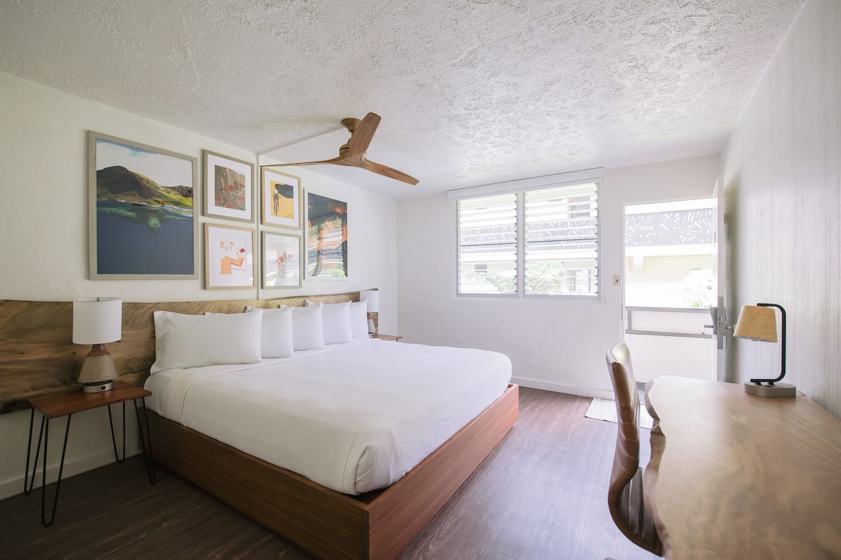 Quarto com cama de madeira, escrivaninha, cadeira, ventoinha no teto e quadros nas paredes.