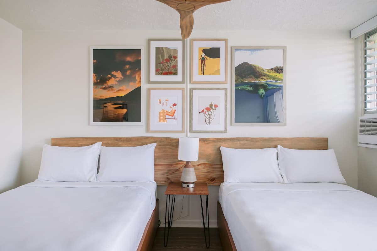 Quarto de hotel com duas camas, cabeceira de madeira, mesa de cabeceira com abajur e quatro quadros na parede.