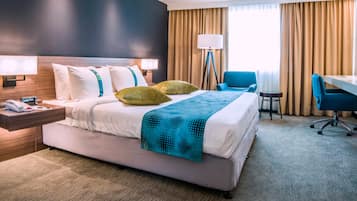 Quarto de hotel com uma cama grande, uma mesa, uma cadeira, um abajur e vista para o exterior.
