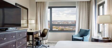 Quarto de hotel com uma cama grande, uma escrivaninha com cadeira, televisão e vista para a cidade através de uma janela ampla.