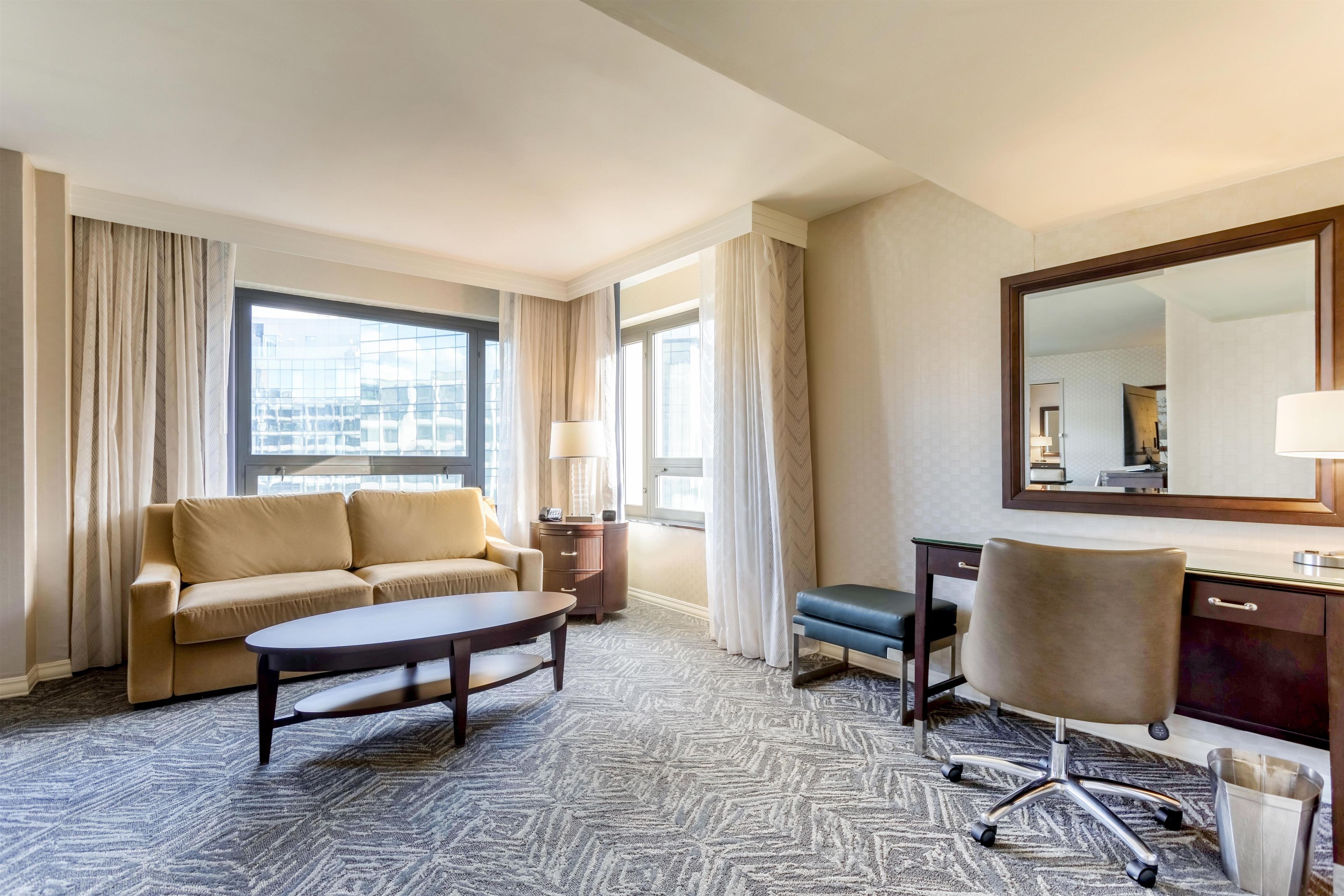 Quarto de hotel com sofá, escrivaninha, cadeira, mesa pequena, abajur e espelho grande.