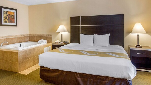 Quarto de hotel com uma cama grande, banheiro separado com banheira, duas luminárias de cabeceira e um quadro na parede.