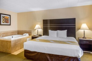 Quarto de hotel com uma cama grande, banheiro separado com banheira, duas luminárias de cabeceira e um quadro na parede.