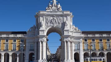 Uma imponente arcada com esculturas detalhadas e uma estátua no topo, ladeada por edifícios com varandas e janelas em arco.
