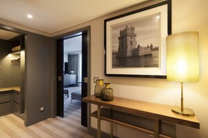 Corredor moderno de hotel com fotografia de um castelo emoldurada, mesa consola de madeira com vasos decorativos e uma luminária.