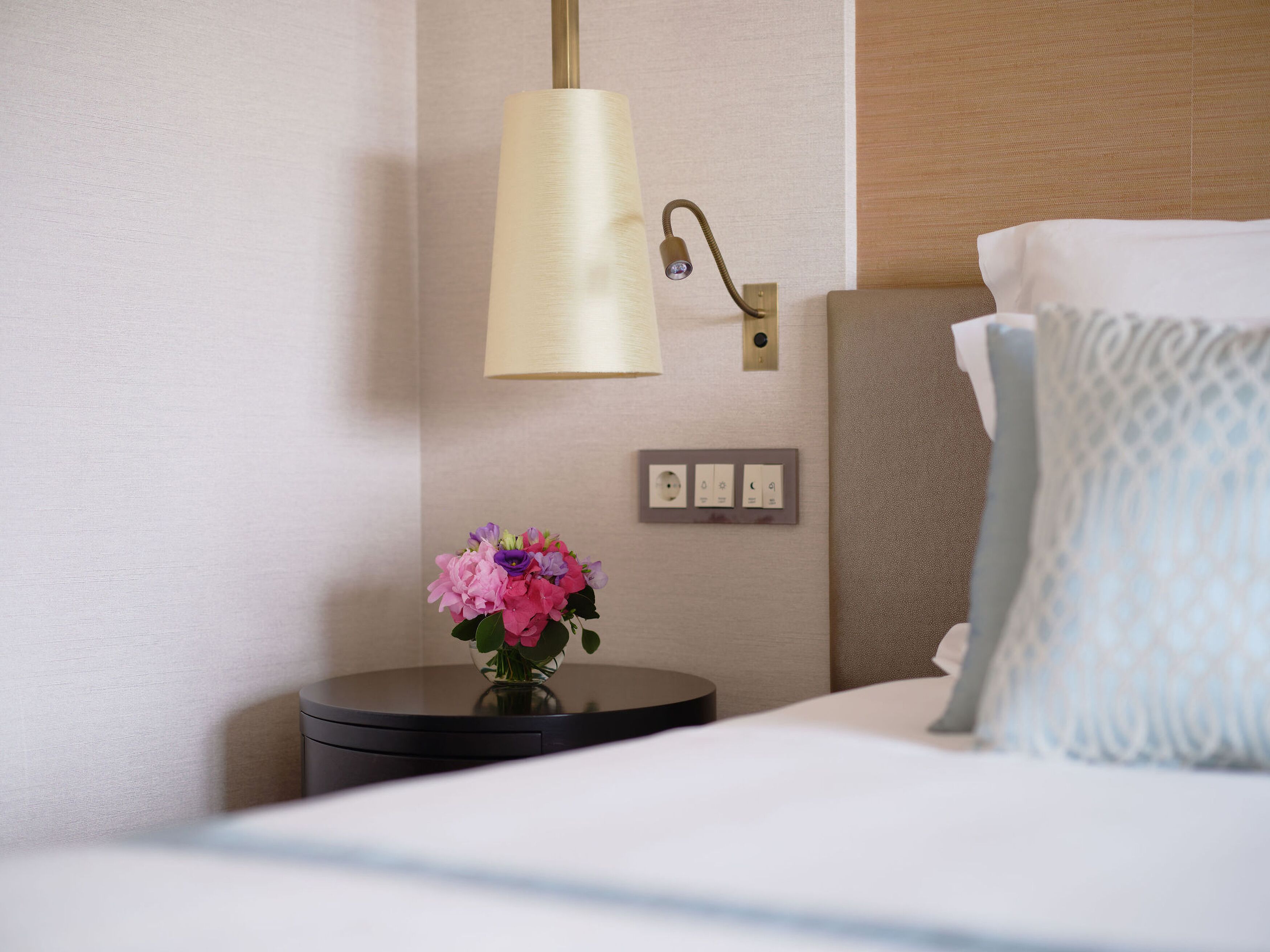 Quarto de hotel com cama, mesa de cabeceira com um vaso de flores, luminária na parede e tomada.