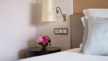 Quarto de hotel com cama, mesa de cabeceira com um vaso de flores, luminária na parede e tomada.