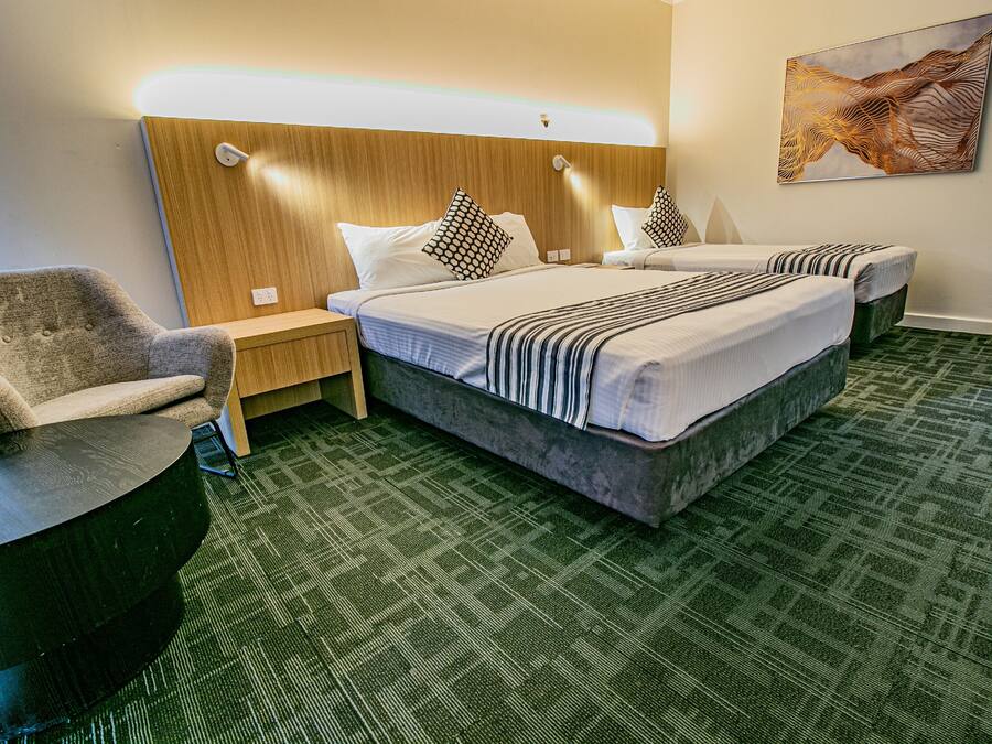 Quarto de hotel com uma cama grande, duas mesas de cabeceira, uma cadeira e um tapete verde.