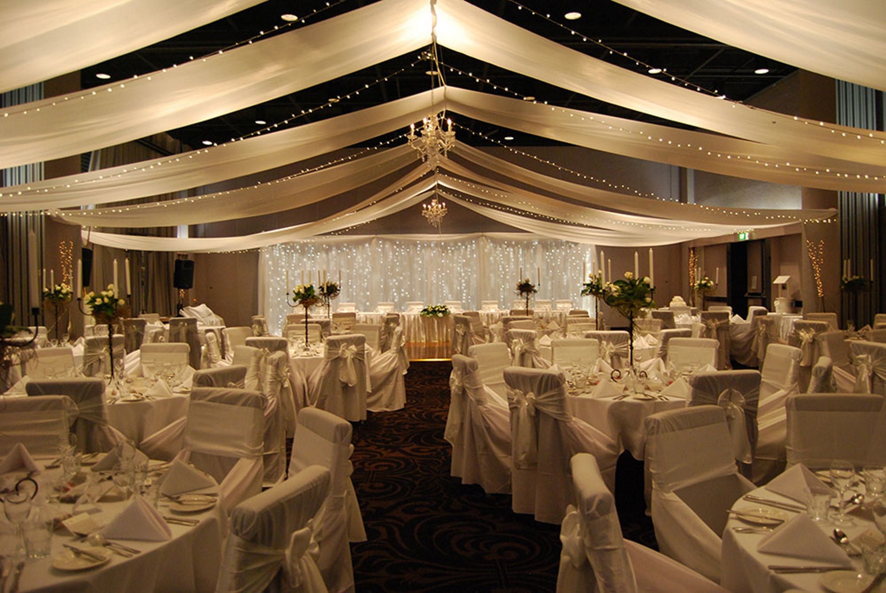 Salão de banquetes com mesas montadas para um evento formal, decorado com tecidos brancos e lustres.