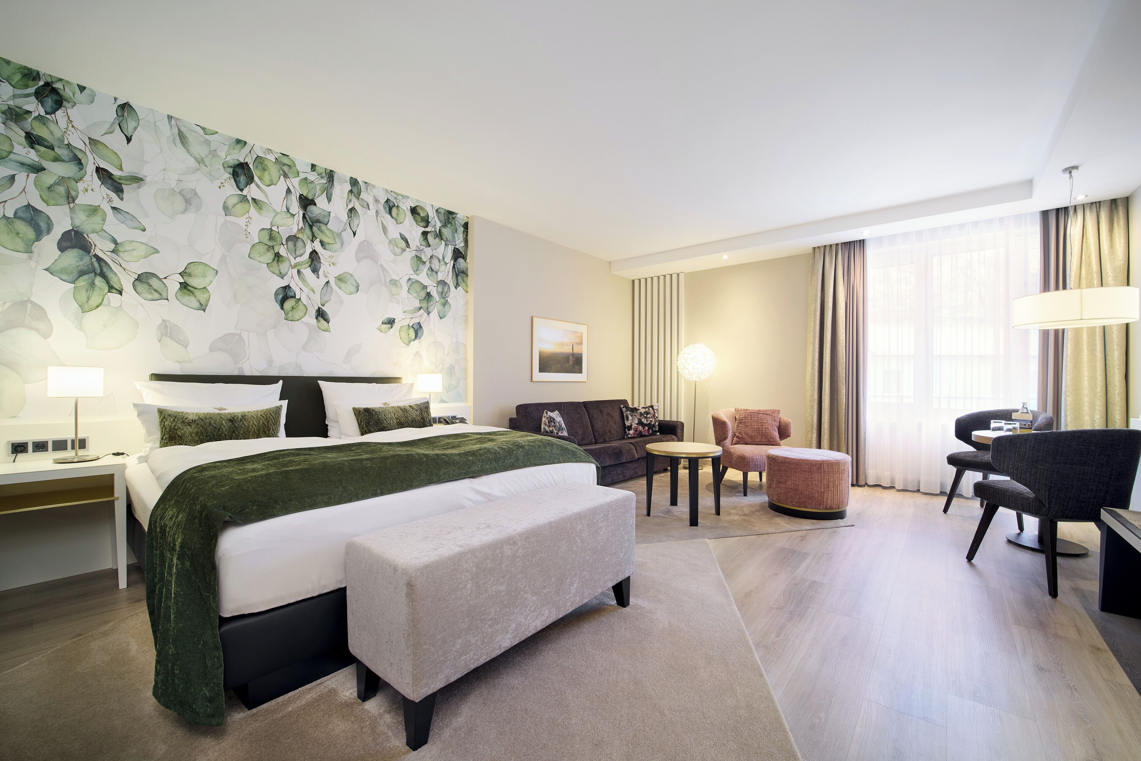 Quarto de hotel com cama, escrivaninha, cadeira, sofá e parede com padrão de folhas.