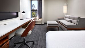 Quarto de hotel com televisão de tela grande, escrivaninha com cadeira, sofá, apoio para os pés e abajur.