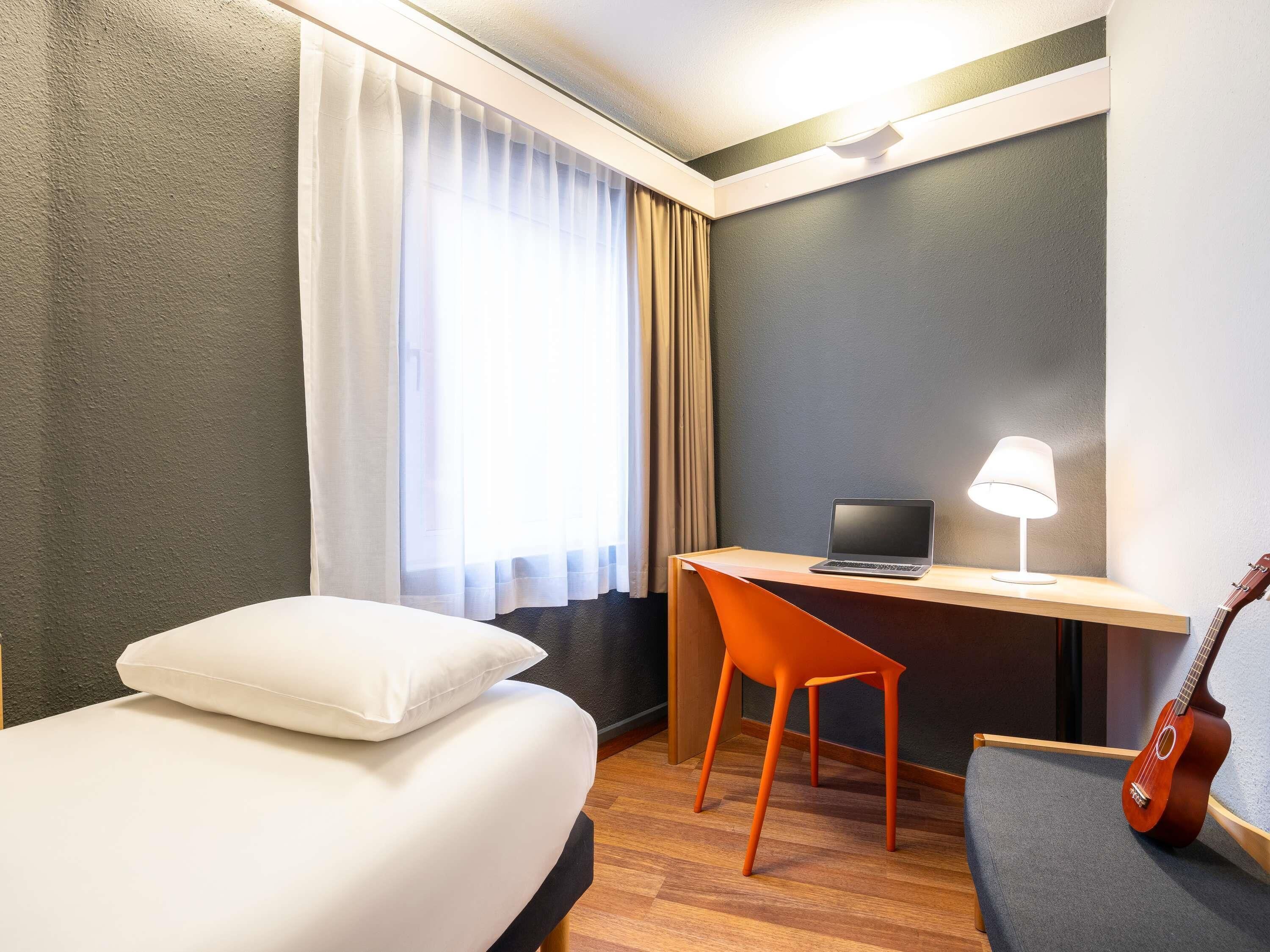 Quarto de hotel com cama, escrivaninha com laptop, cadeira, guitarra e luminária.