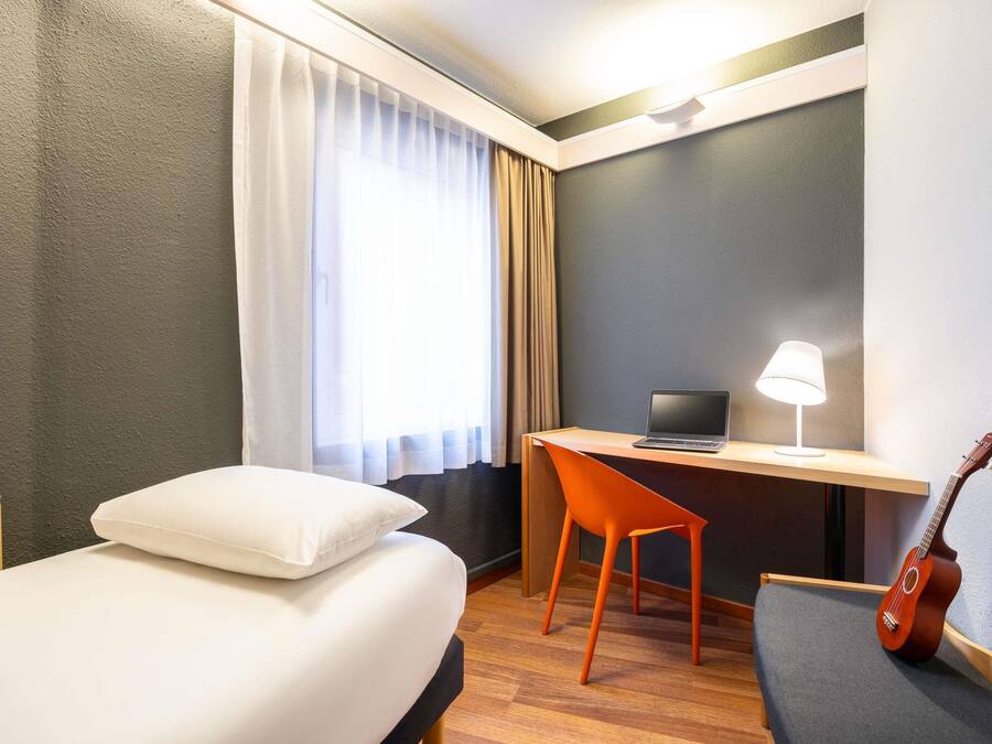 Quarto de hotel com cama, escrivaninha com laptop, cadeira, guitarra e luminária.