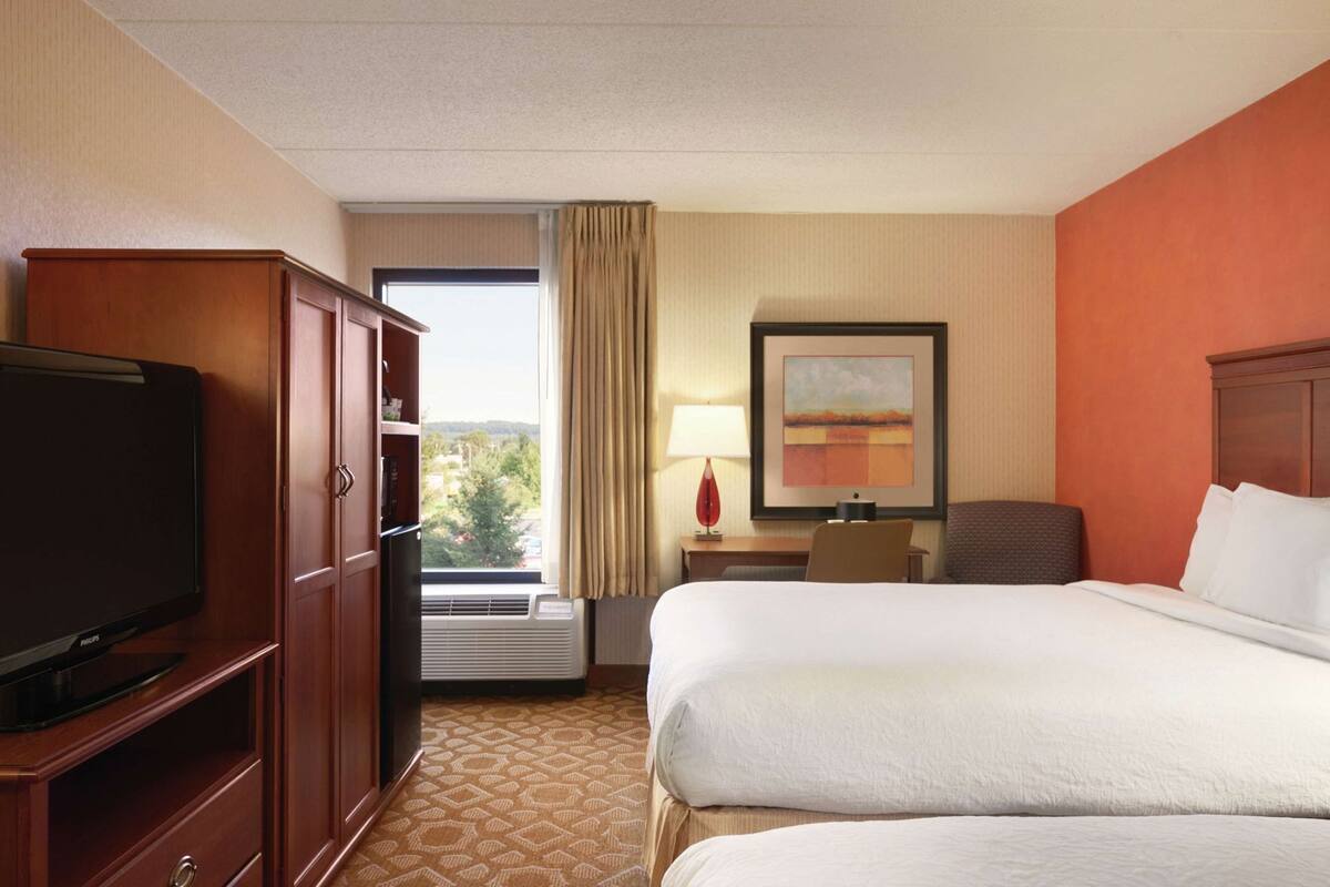 Quarto de hotel com cama, televisão, escrivaninha com luminária e janela com cortinas.