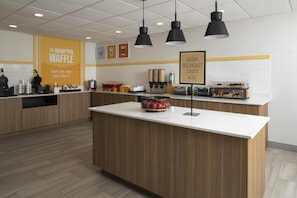 Uma cozinha moderna com uma ilha central, armários de madeira e um balcão para refeições rápidas. Há waffle makers, máquinas de café e uma cesta de frutas no balcão.