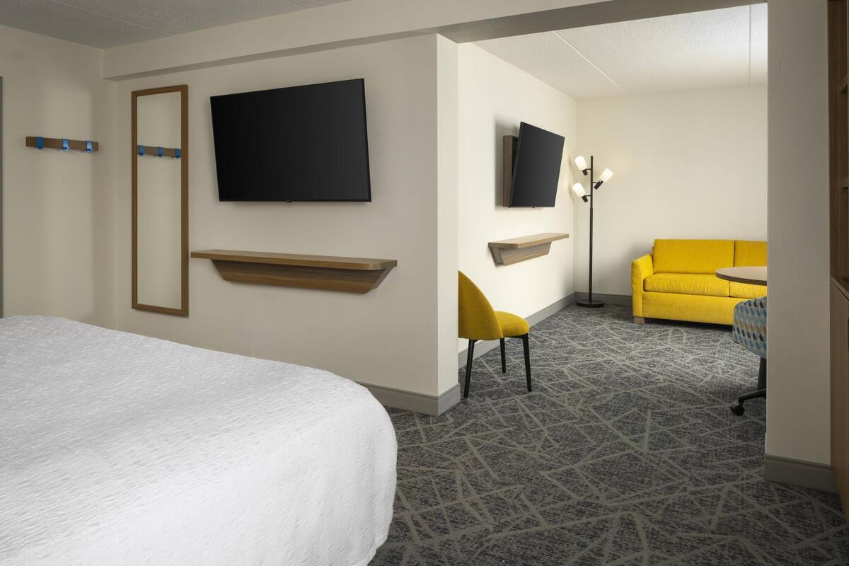 Quarto de hotel com uma cama grande, televisão de tela plana na parede, uma escrivaninha pequena com cadeira, um sofá amarelo e um abajur.