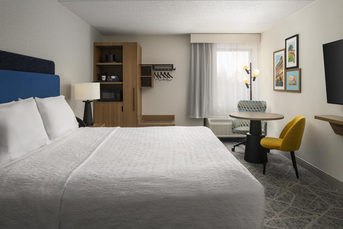 Quarto de hotel com uma cama grande, uma escrivaninha com cadeira, um guarda-roupa, uma mesa pequena e uma televisão na parede.