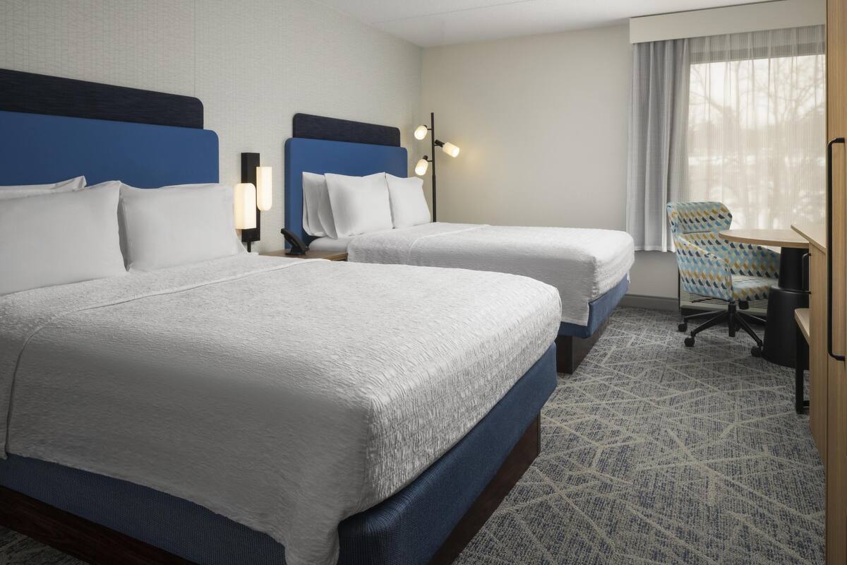 Quarto de hotel com duas camas, uma escrivaninha, uma cadeira e uma janela grande com cortinas.