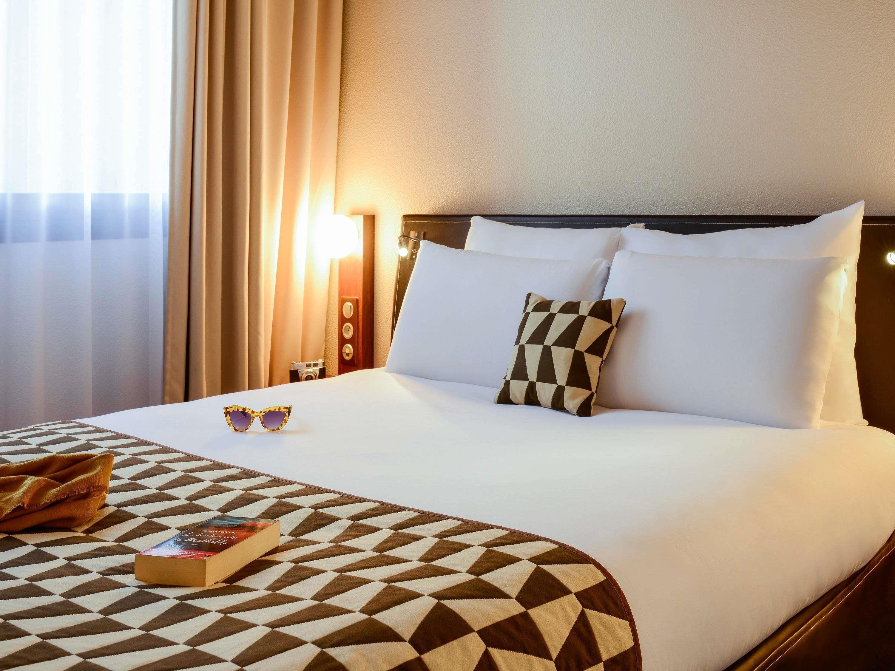 Quarto de hotel com uma cama, manta estampada, um livro e um par de óculos de sol sobre a cama.