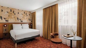 Quarto de hotel com uma cama, uma cadeira, uma mesa pequena e um detalhe decorativo na parede.