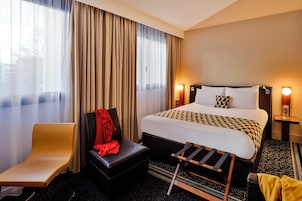 Quarto de hotel com cama, uma cadeira, uma mesinha e janela com cortinas.