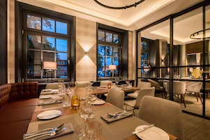 Uma sala de jantar bem iluminada com uma mesa comprida posta para vários convidados, grandes janelas com cortinas e vista para o exterior.