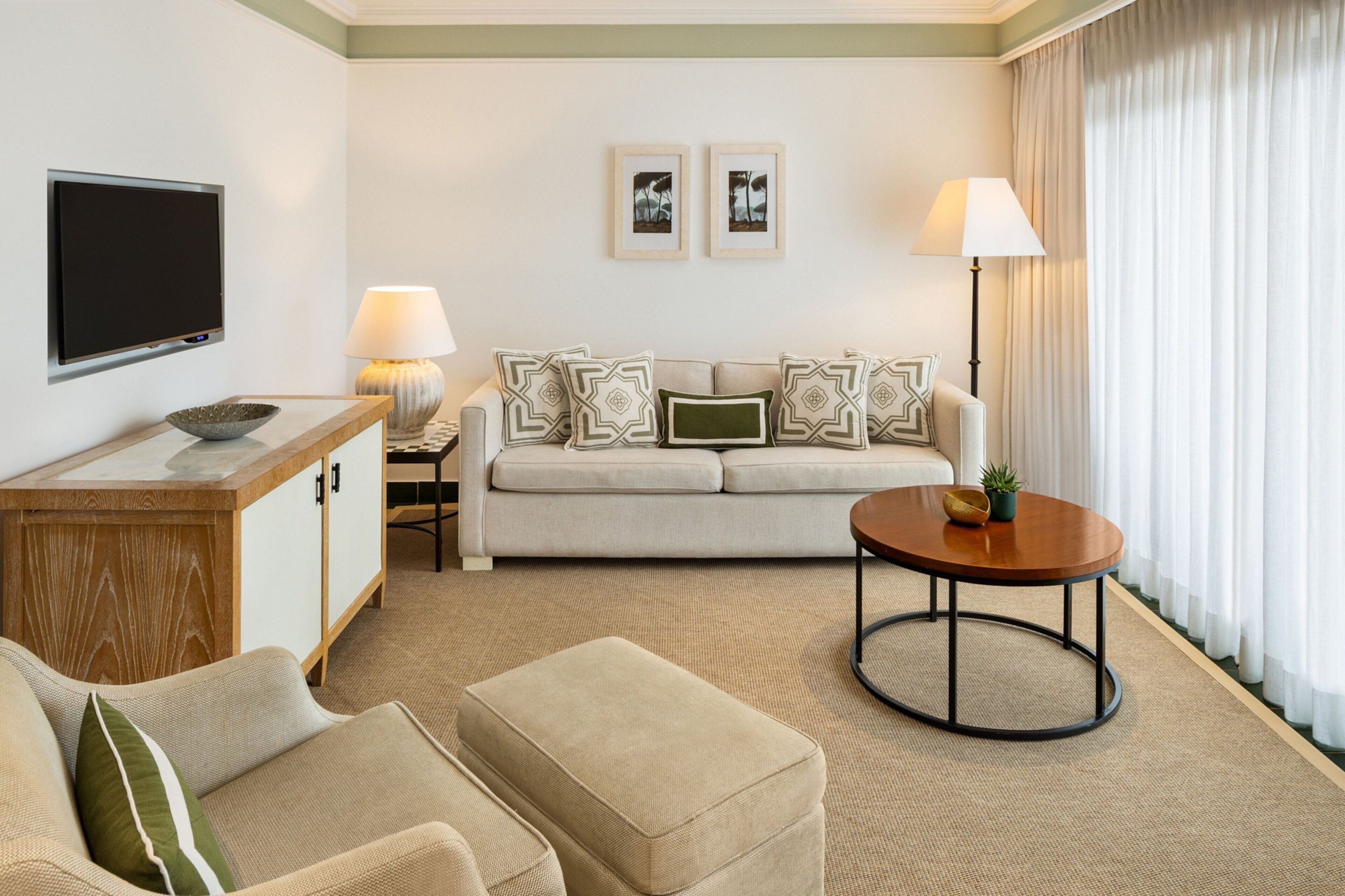 Quarto de hotel moderno com sofá, duas poltronas, mesa de centro, TV e escrivaninha com abajur.