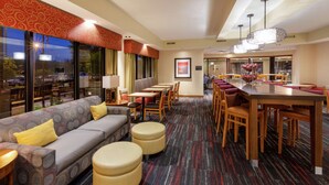 Um hall de hotel com um sofá, pufes, uma área de refeições com uma mesa comprida e cadeiras, e grandes janelas com vista para a rua.