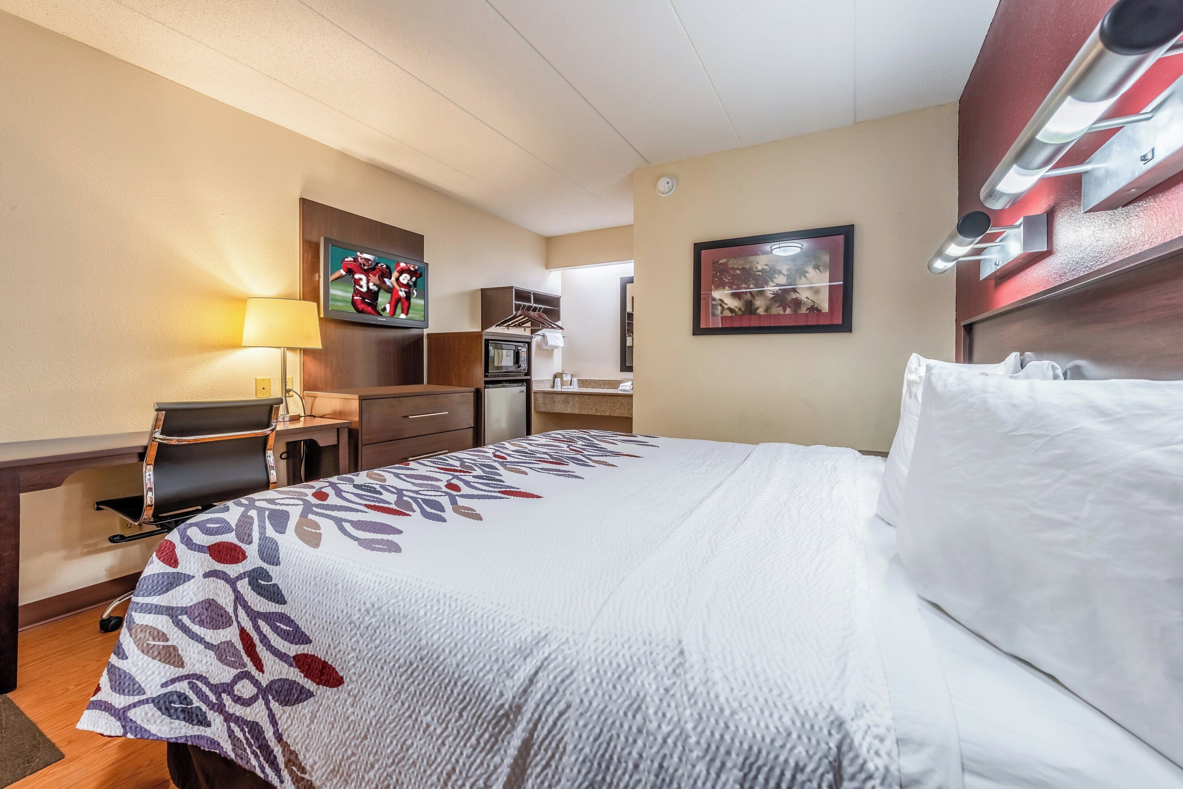 Quarto de hotel com cama, escrivaninha, televisão, micro-ondas e quadro na parede.