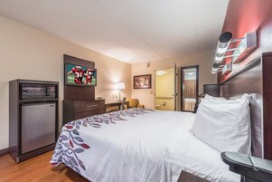 Quarto de hotel com cama, escrivaninha, micro-ondas, televisão e porta para o banheiro.