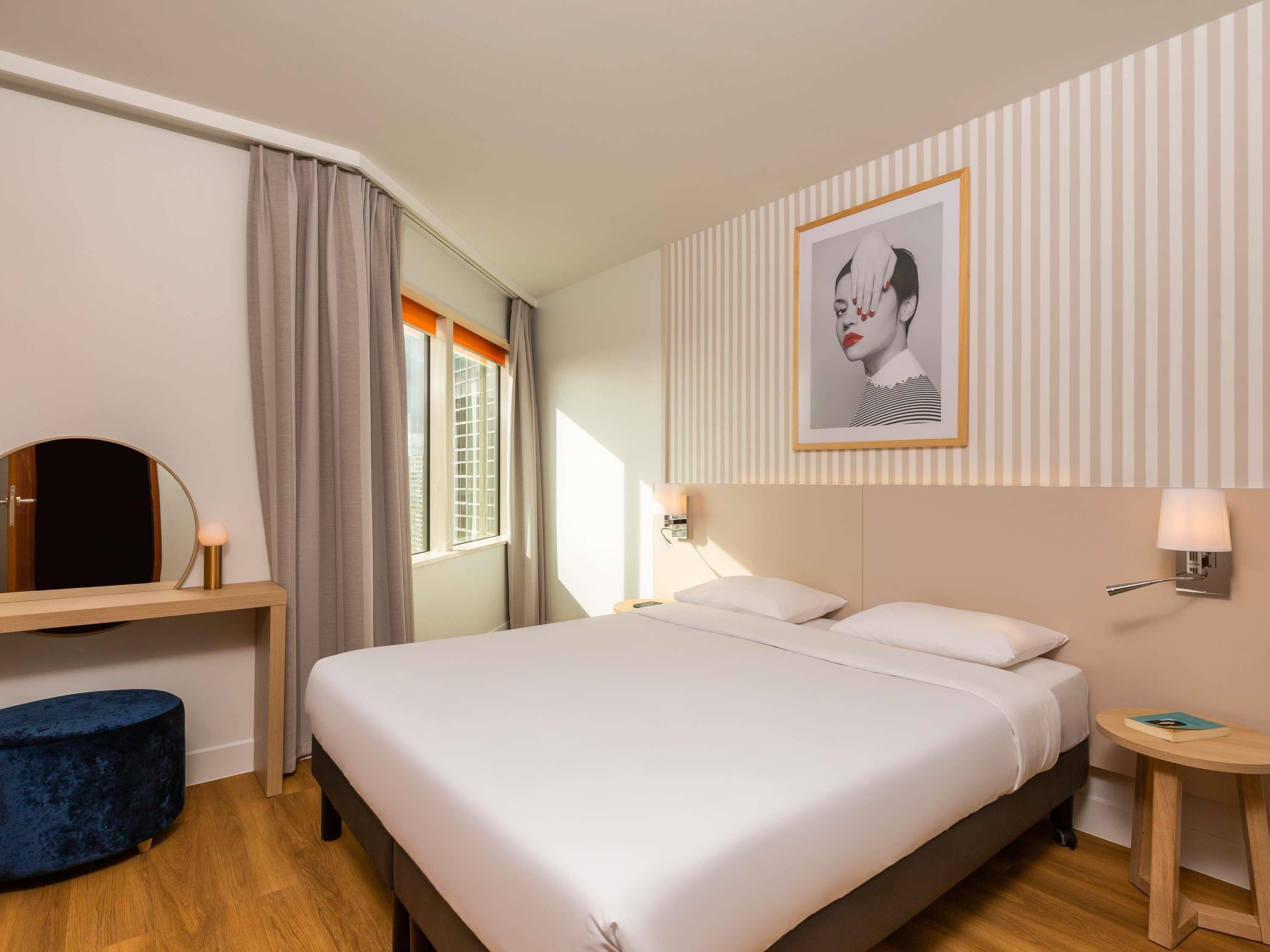 Quarto de hotel com cama, mesas de cabeceira, espelho, janela com cortinas, um quadro pendurado na parede e uma pequena mesa redonda.