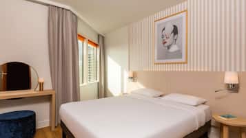 Quarto de hotel com cama, mesas de cabeceira, espelho, janela com cortinas, um quadro pendurado na parede e uma pequena mesa redonda.