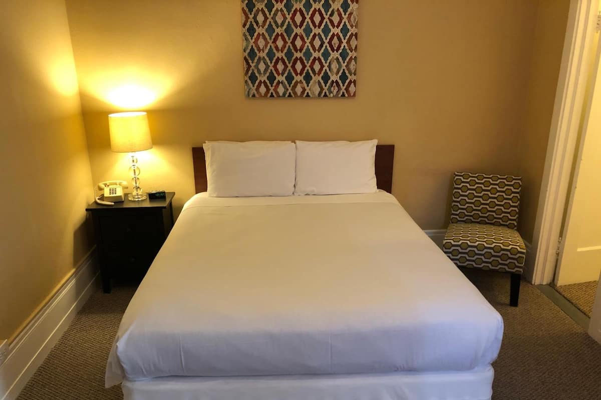 Quarto de hotel com uma cama grande, uma mesinha de cabeceira com luminária, um telefone e uma cadeira estampada.