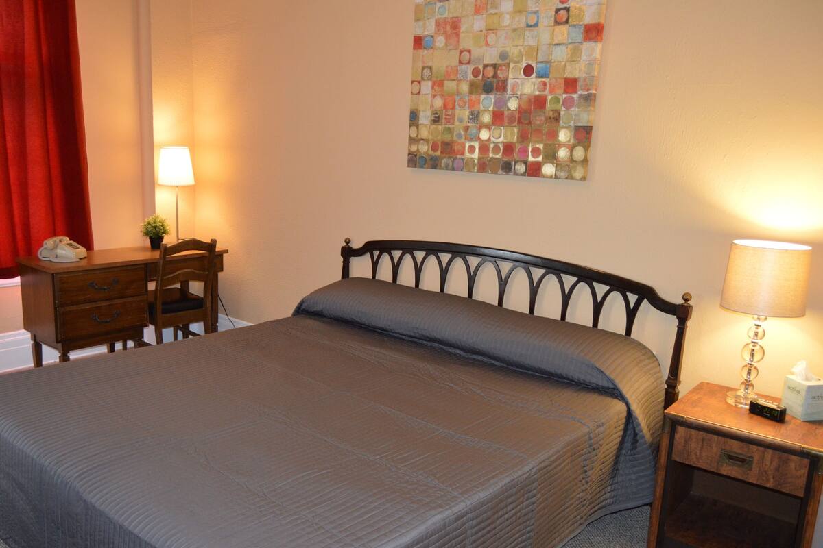 Quarto com cama, escrivaninha, cadeira, mesa de cabeceira, abajur e uma obra de arte na parede.