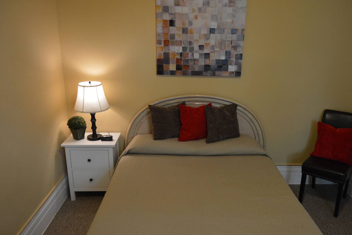 Quarto bem arrumado com cama, mesa de cabeceira, abajur e cadeira.
